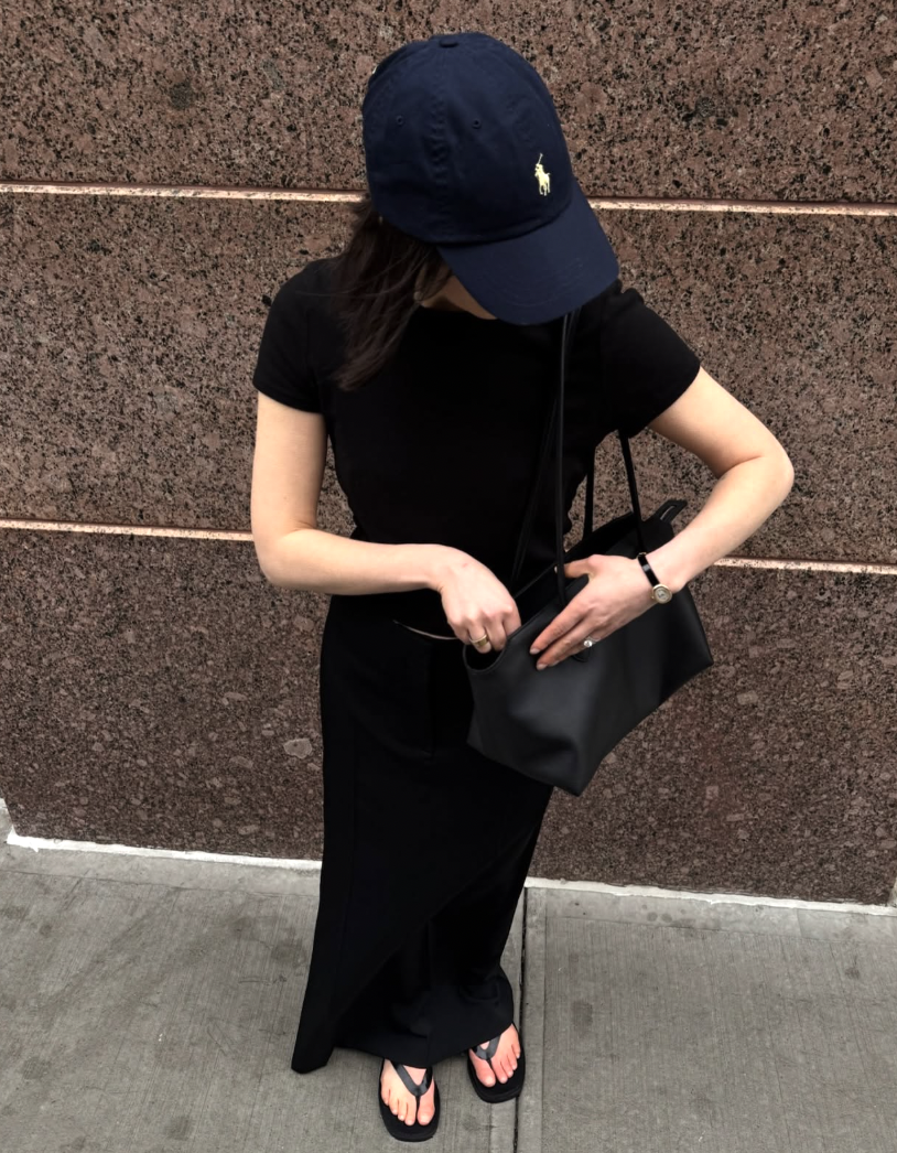 Woman wearing a black fitted t-shirt dress with a navy blue Polo Ralph Lauren cap%2C black leather shoulder bag%2C and black flip-flops on a city sidewalk